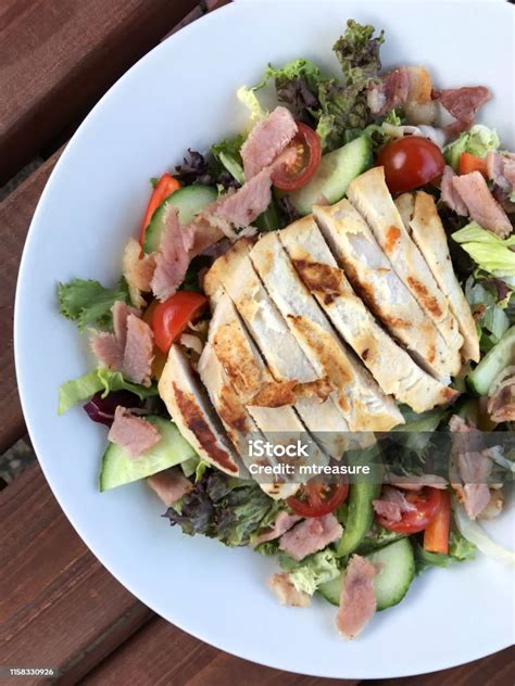 바삭바삭한 베이컨 혼합 샐러드 양상추 잎 슬라이스 양파 체리 토마토 오이 로켓 피망과 샐러드 드레싱 나무 피크닉 테이블과 치킨 시저 샐러드 조리법의 야외 술집 Grub을