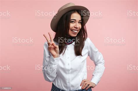명랑 세련 된 소녀의 초상화 V 기호 또는 평화 제스처를 보여줍니다 분홍색 배경 위에 고립 된 흰색 셔츠와 모자를 착용 매우