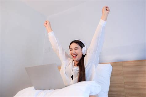 Asian Women Sitting On Bed In Bedroom Wearing Headphone Looking Down On Laptop Cheer For Her