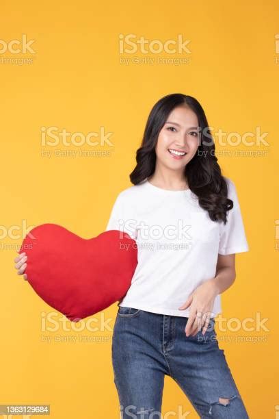 행복한 아시아 젊은 여성은 노란색 배경에 고립 된 귀엽고 사랑스러운 미소 빨간 마음을 들고 긴 곱슬 머리 사랑과 발렌타인 데이