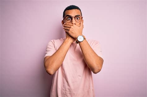 캐주얼 한 티셔츠와 안경을 분홍색 배경 위에 입은 잘 생긴 아프리카 계 미국인 남자는 실수로 손으로 입을 덮는 충격을 받았습니다