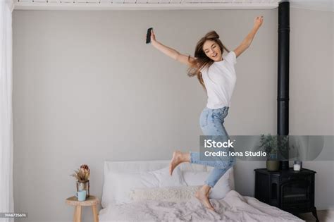 흰색 티셔츠와 파란색 데님 청바지를 입은 젊은 성인 이탈리아 여성이 집에서 침대에서 행복한 점프를 하고 있다 백인 소녀를 축하하는 복사 공간이 있는 모형 행복과 젊음 30