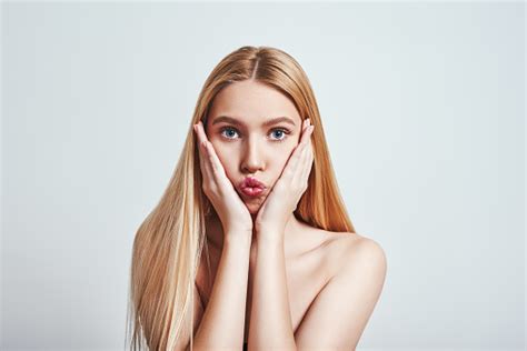 제가 뭘 해야 하나요 긴 금발 머리를 가진 젊은 여자는 카메라를 보고 하 고 회색 배경에 서 있는 동안 얼굴을 만들기 귀여운에