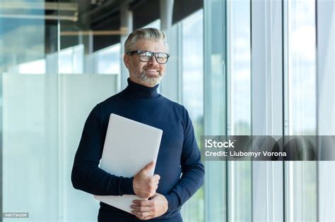 광범위한 업무 경험을 가진 오래된 디자이너 건축가 엔지니어의 초상화 그는 노트북을 손에 들고 창가에 사무실에 서 있습니다 자신