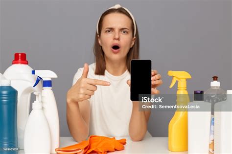 충격을 받고 놀란 놀란 여자는 회색 배경 위에 격리된 청소 세제를 들고 직장에서 포즈를 취하며 광고를 위한 빈 공간을 가리켰다
