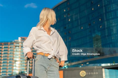 초상화 젊은 금발의 여자는 설정 태양의 광선에 현대적인 유리 호텔의 배경에 그녀의 손에 가방과 흰색 셔츠와 청바지에 카메라에서