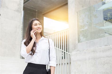 아름 다운 매력적인 아시아 비즈니스 여자 미소 너무 행복을 느끼고 그녀의 직업과 요구를 사무실 밖에 서 스마트폰에서 고객과 얘기 하 고 자신감 비즈니스 개념 거리에 대한