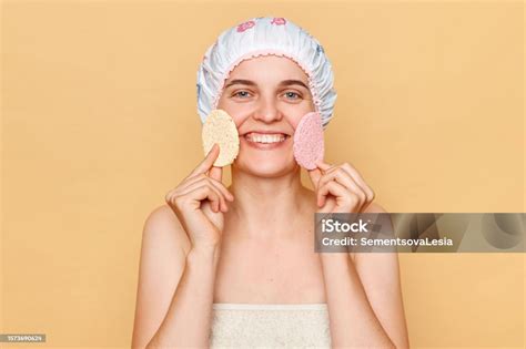 피부 관리 미용 및 얼굴 치료 스펀지를 들고 웃는 아름다운 여자는 목욕 후 피부가 매끄럽고 샤워 캡과 수건을 쓰고 베이지색 배경에 서 있다 목욕 스폰지에 대한 스톡 사진 및
