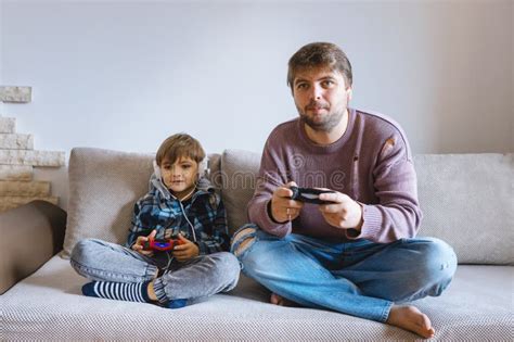 Father And Son Sitting On Sofa In Lounge Playing Video Game Stock Photo