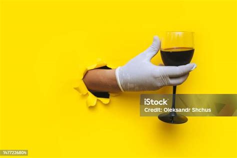 밝은 노란색 종이의 찢어진 구멍에서 오른쪽 의사의 손이 적포도주 한 잔과 함께 흰색 라텍스 장갑에 나타납니다 알코올 술 취함 및