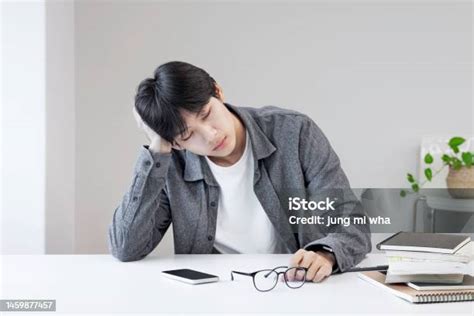 책상 위에 책과 휴대폰을 들고 있는 현대적인 홈 오피스에서 두통으로 고통받는 젊은 아시아 대학생 남성 편두통을 느끼는 과로 한국 학생들 얼굴을 손에 기댐에 대한 스톡 사진