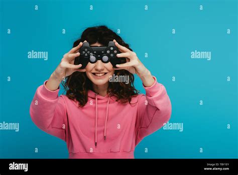 Cheerful Curly Redhead Girl Covering Face With Joystick On Blue Stock