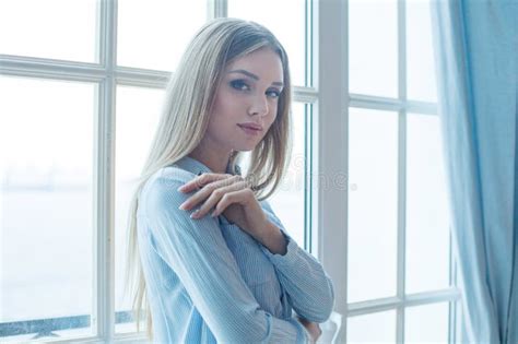 очаровательная молодая женщина с длинными светлыми волосами и макияжом позирующая перед окном