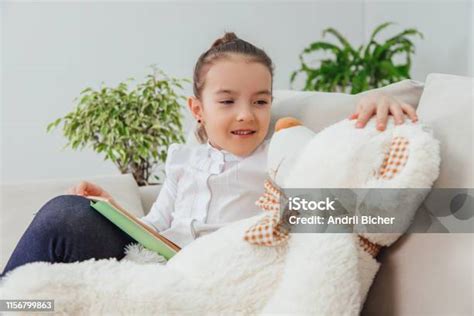 귀여운 어린 소녀는 그녀의 근처에 테디 베어와 소파에 앉아 동화를 읽고 머리에 그녀의 부드러운 친구를 애무 땋은 머리에 대한