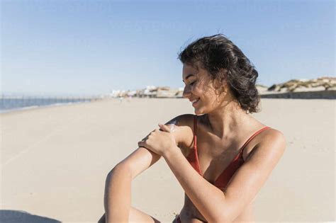 Smiling Young Woman Applying Sunscreen While Sitting At Beach On Sunny