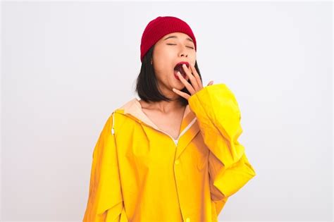 고립 된 흰색 배경 위에 비옷과 양모 모자를 쓰고 있는 젊은 아름다운 중국 여성은 손으로 입을 덮는 피곤한 하품 불안과 졸음 가리기에 대한 스톡 사진 및 기타 이미지