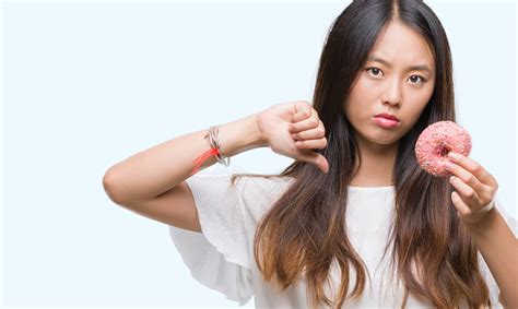 성 난 얼굴 아래로 엄지 싫어 보여주는 음수 부호 거부 개념 고립 된 배경 위에 도넛을 먹는 젊은 아시아 여성 개념에 대한 스톡