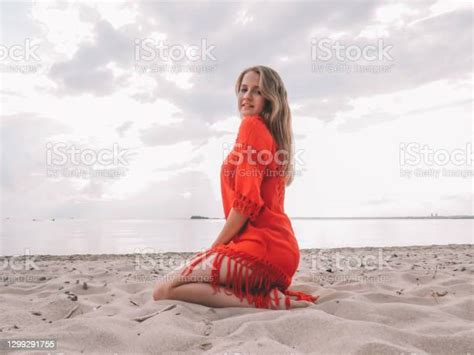 빨간 여름 드레스에 긴 금발 머리와 슬림 매력적인 몸을 가진 무두질 젊은 여성은 물과 그림 같은 하늘의 배경에 모래 해변에서 포즈 18 19세에 대한 스톡 사진 및 기타