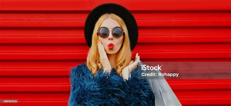 파란 인조 모피 코트를 입고 쇼핑백을 들고 충격을 받은 젊은 여성 빨간 벽 배경 위에 검은 둥근 모자를 들고 있는 충격적인 젊은 여성 25 29세에 대한 스톡 사진 및 기타