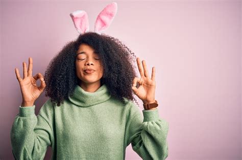 분홍색 배경 위에 부활절 토끼 귀 의상을 입고 아프리카 계 미국인 여성이 손가락으로 명상 제스처를하고 눈을 감고 미소 요가