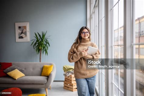 식물과 화려한 쿠션으로 가득한 아늑하고 현대적인 생활 공간에서 큰 창문 근처에 서서 태블릿을 들고 있는 젊은 여성 가구에 대한 스톡 사진 및 기타 이미지 Istock