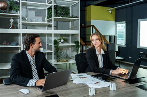 사무실 책상에서 따로 일하는 백인 사업가와 사업가는 둘 다 현대적인 작업 공간에서 노트북을 사용하고 프린트 차트가 테이블 위에 놓여 있고 정장을 입고 캐주얼하게 시선을