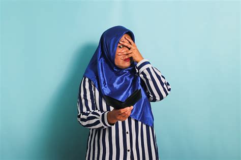 히잡을 쓴 충격적인 중년 아시아 여성이 파란색 배경에 고립된 빈 지갑을 들여다보고 있다 50 54세에 대한 스톡 사진 및 기타