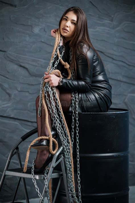 A Striking Young Woman With Long Dark Hair Sits On A Black Barrel Bound With Rope And Chains