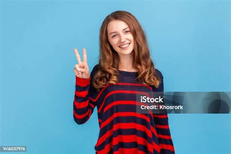손가락으로 평화의 상징을 보여주는 줄무늬 스웨터를 착용하고 행복한 만족한 여성 이빨 미소로 카메라를 바라보며 승리를 축하합니다