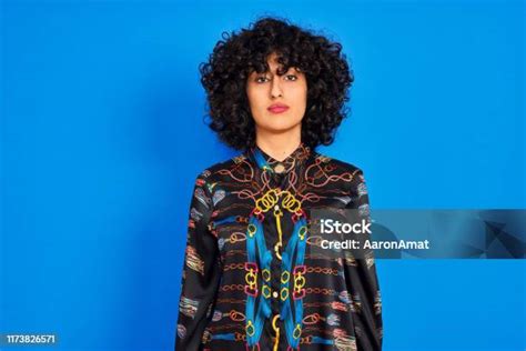 얼굴에 심각한 표정과 고립 된 파란색 배경 에 화려한 셔츠를 입고 곱슬 머리를 가진 젊은 아랍 여성 카메라를 보는 간단하고 자연스러운 결심에 대한 스톡 사진 및 기타 이미지