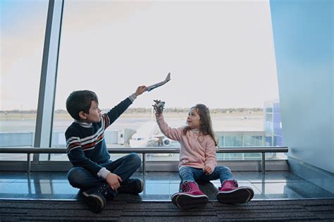 활주로가 내려다 보이는 공항 출발 터미널의 파노라마 창문 옆에 바닥에 앉아있는 장난감 비행기를 가지고 노는 어린 소년과 소녀