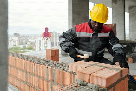 미완성 건축물 안에 서 있는 동안 특수 망치로 벽돌 층의 표면을 평평하게 하는 작업복의 숙련된 건축업자 사진 Unsplash의 노동 이미지