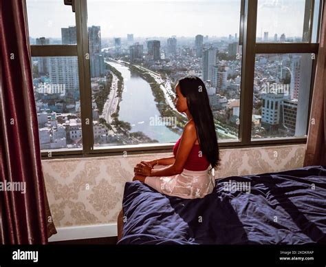 Side View Of An Asian Woman Sitting On A Bed At The Window With The