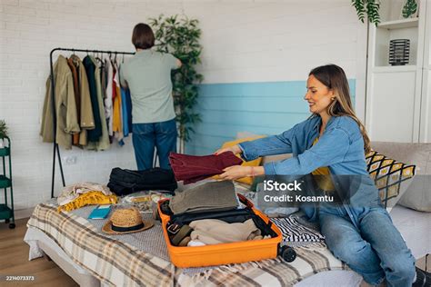 한 남자가 옷걸이 앞에 서서 재킷과 상의로 여행을 위해 무엇을 챙길지 고르고 있습니다 그의 여자친구는 침대에 앉아서 여행을 위한 여행 가방을 챙기고 있습니다 2명에 대한