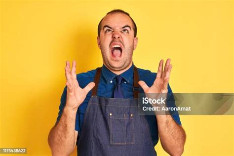 고립된 노란 배경 위에 앞치마를 입고 있는 젊은 가게 주인 남자는 공격적인 표정과 팔을 들고 미친 듯이 외치고 소리쳤다 좌절 개념입니다 분노에 대한 스톡 사진 및 기타