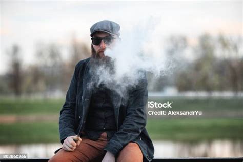 Vape 실제 생활에서 남자 수염 봄 저녁에 강 근처 제방에서 증기의 퍼프와 전자 담배는 모자와 선글라스 Vaping에 큰