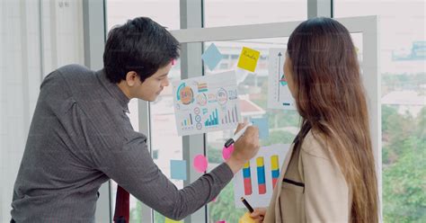 비즈니스 사람들은 사무실의 프로젝트 계획 보드에서 일하고 동료 친구와 대화하여 프로젝트 개발을 분석합니다 그들은 유리 벽에