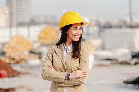 迷人的女建筑师棕色头发，牙齿的微笑，戴上头盔，穿着时髦的休闲站在建筑工地与双臂交叉。