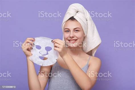 손에 얼굴 마스크를 가진 소녀의 초상화를 닫고 회색 티셔츠를 입고 머리에 수건을 쓰고 라일락 배경으로 고립 된 포즈를 취합니다 아름다운 소녀는 그녀의 얼굴에 화장품 마스크를