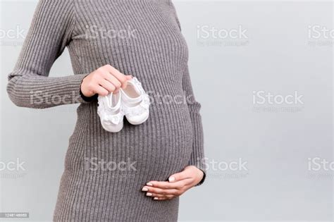 회색 배경에 임신 한 여성의 손에 아기 소녀를위한 신발의 클로즈업 미래의 어머니는 회색 드레스를 입고 있습니다 모성 개념 복사
