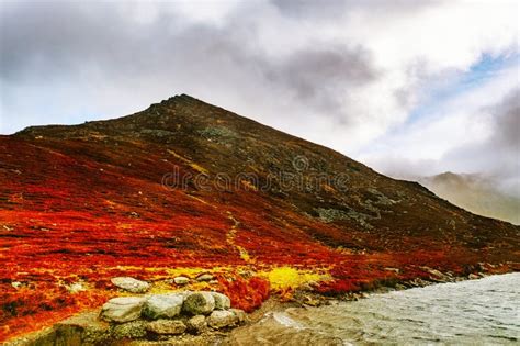 A Grassy Hillside Slopes Up Towards A Rocky Peak Under A Cloudy Sky A Small Body Of Water