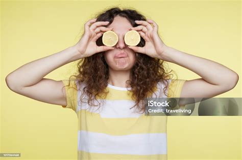 곱슬 머리 재미 여자 닫습니다 레몬의 2 개 반으로 눈 및 입을 노란색 배경 위에 고립 된 과일의 신 맛을 보여주는 혀를 보여