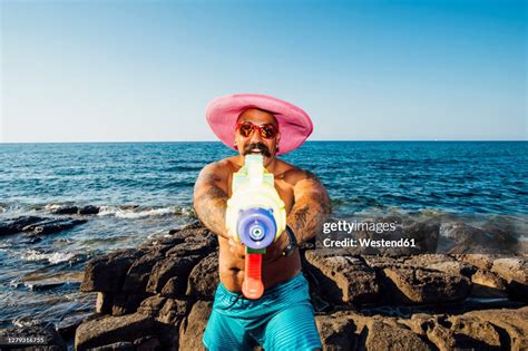 Man Wearing Hat Showing Squirt Gun While Standing Against Sea High Res