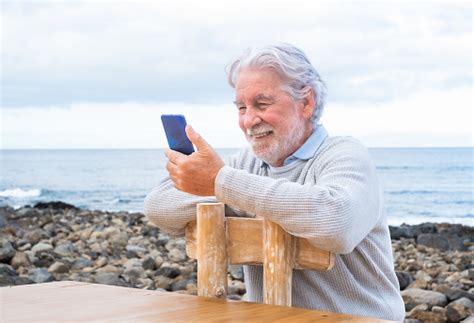 수염을 가진 편안한 노인이 휴대 전화를 사용하여 바다에서 야외에 앉아 웃고 있습니다 흰 머리의 백인 노인 할아버지가 스마트 폰을 들고있는 나무 의자에 해변에 앉아 있습니다