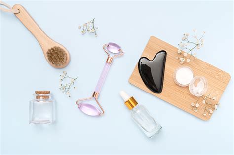 핑크 석영 스톤 롤러 얼굴 마사지기 구아슈 파스텔 블루 배경에 유리 화장품 병과 흰색 꽃 홈 스파 케어 아름다움과 젊음 안티