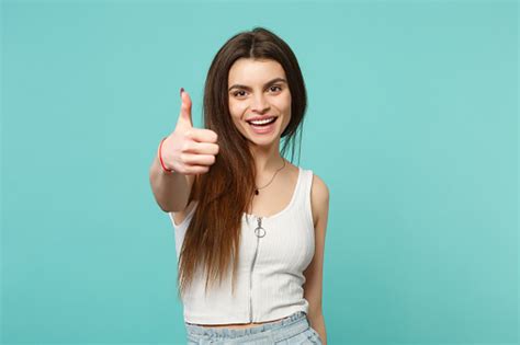 스튜디오에서 파란색 청록색 벽 배경에 고립 된 엄지손가락을 보여주는 가벼운 캐주얼 복장에 웃는 젊은 여자의 초상화 사람들은