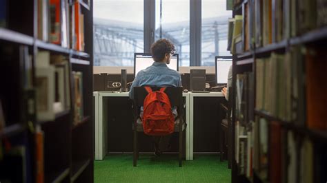 대학 도서관 책상에 앉아 Pc에서 일하고 수업 과제에 대한 연구를 하고 과목을 공부하고 논문을 쓰는 데 초점을 맞춘 학생 책장의