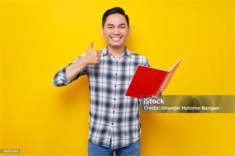 하얀 체크무늬 셔츠를 입고 책을 들고 엄지손가락을 치켜들고 있는 잘생긴 젊은 아시아 남성이 노란색 배경 위에 고립되어 있다 사람들의 라이프스타일 컨셉 30 34세에 대한