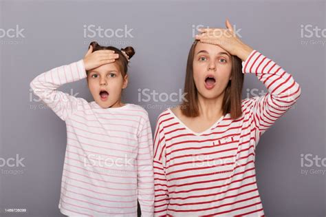 겁에 질린 충격을 받은 여성과 캐주얼한 옷을 입은 어린 소녀가 회색 배경 위에 고립되어 서서 겁에 질린 표정으로 카메라를