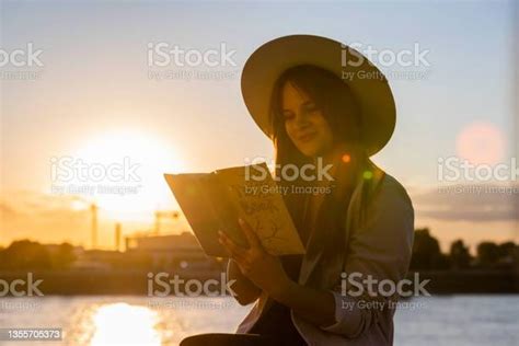 모자를 쓴 젊은 아름다운 여성이 책을 손에 들고 강가에 앉아 있습니다 강과 다리는 배경에 우세합니다 25 29세에 대한 스톡 사진 및 기타 이미지 Istock